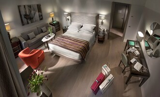 Quarto de hotel moderno com uma cama grande, um sofá, uma escrivaninha e uma cadeira. Há duas mesas de cabeceira com luminárias e uma mesinha com um vaso de flores.