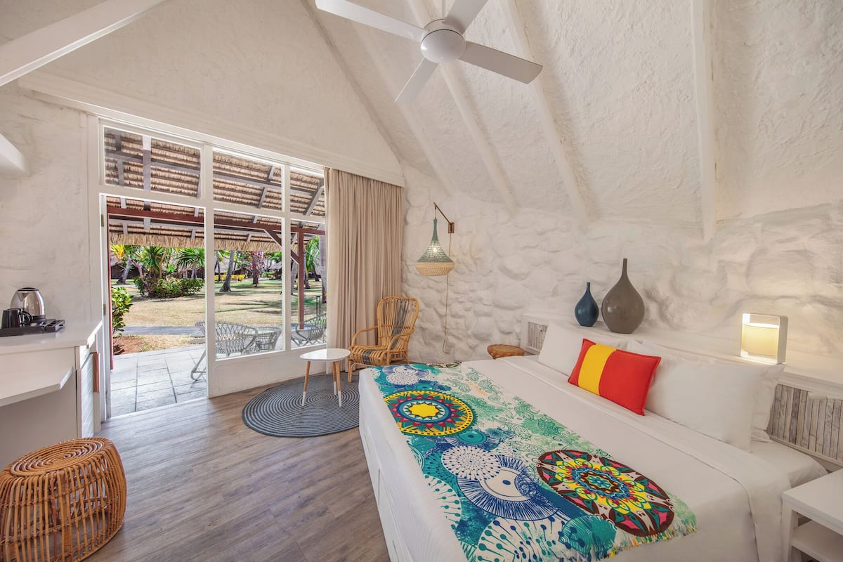 Quarto com cama, escrivaninha e cadeira. Possui uma janela grande com cortinas, ventilador de teto e vista para o exterior.