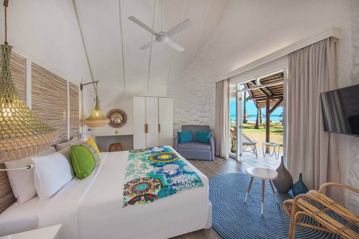 Quarto com cama, ventilador de teto, televisão, varanda com vista para o mar e área de refeições com cadeiras e mesa.