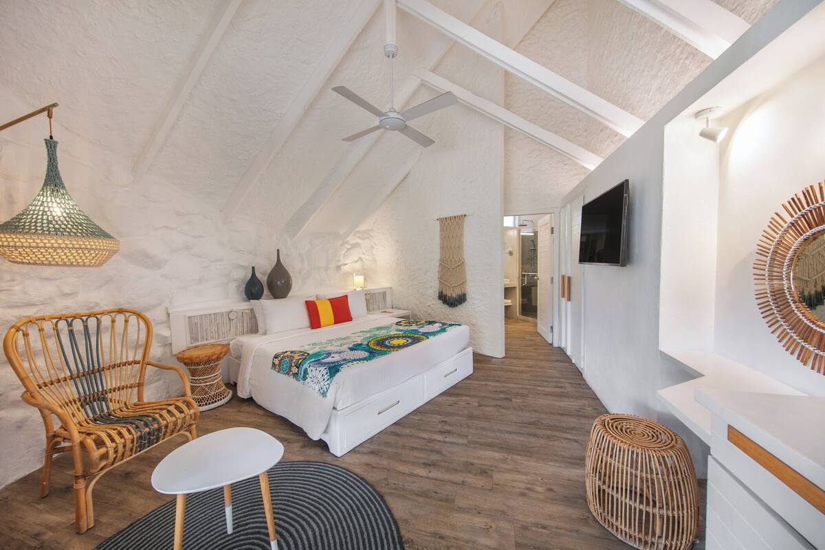 Um quarto com p é-direito alto e teto inclinado, uma cama com manta colorida, uma cadeira de vime, uma pequena mesa e uma televisão fixada na parede.