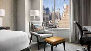 Quarto de hotel com cama, uma cadeira com almofada colorida, uma mesinha, escrivaninha e vista para edifícios da cidade através de uma janela grande.
