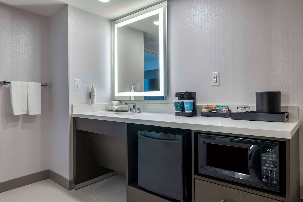 Casa de banho moderna com bancada branca, espelho embutido, máquina de café, micro-ondas e pequeno frigorífico.