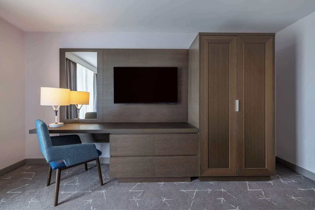 Quarto de hotel com escrivaninha, cadeira, televisão, guarda-roupa e luminária.