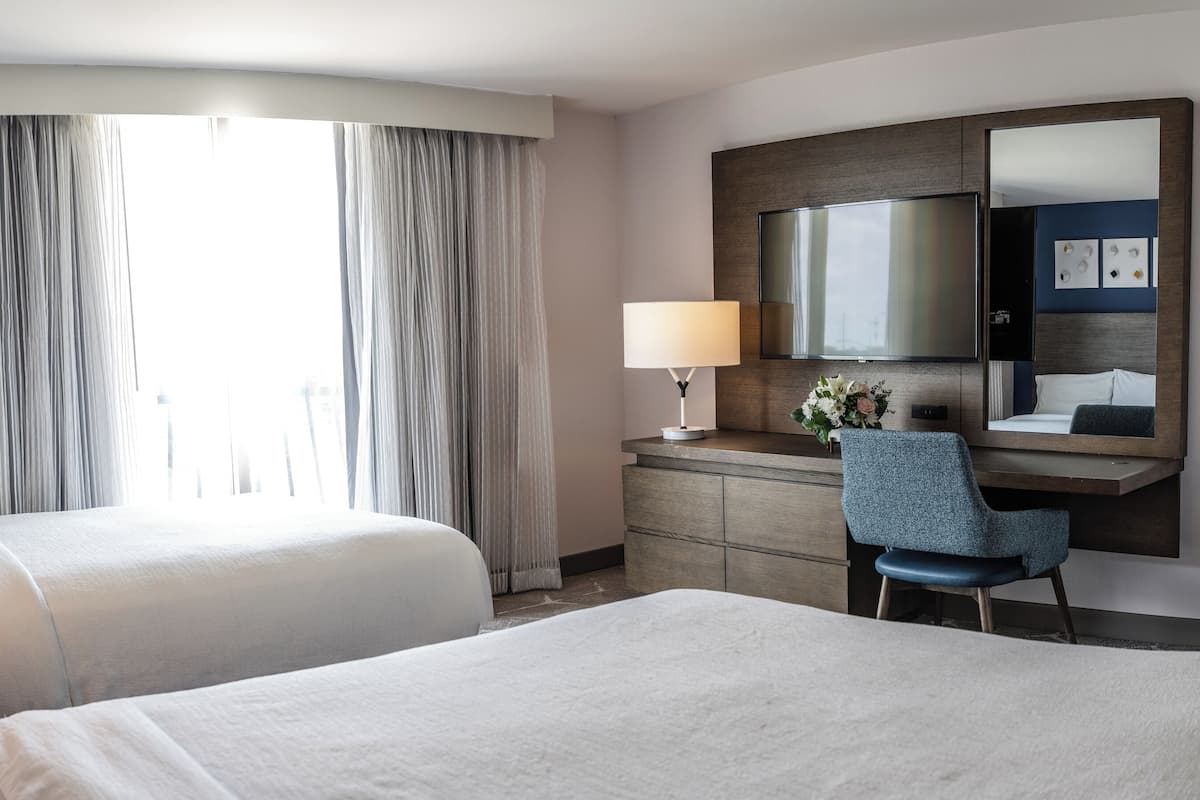 Quarto de hotel com duas camas, uma secretária com cadeira, televisão e uma janela grande com cortinas.