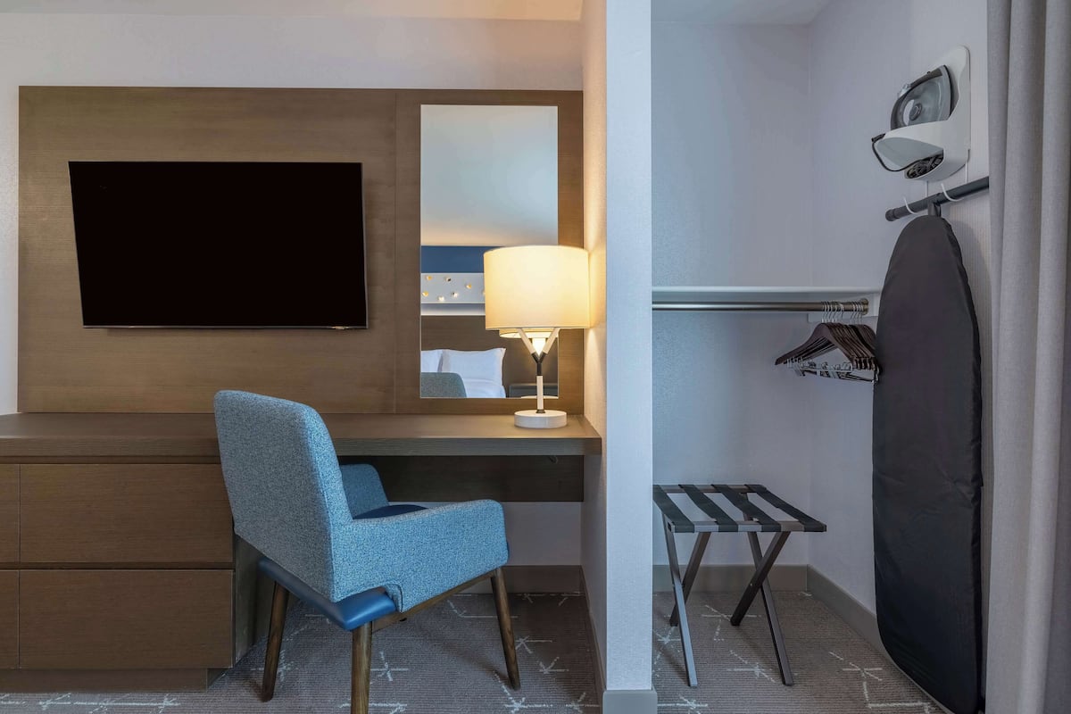 Quarto de hotel com secretária de madeira, uma cadeira azul, televisão de tela plana, abajur, cabideiro e um pequeno ferro de passar roupa.