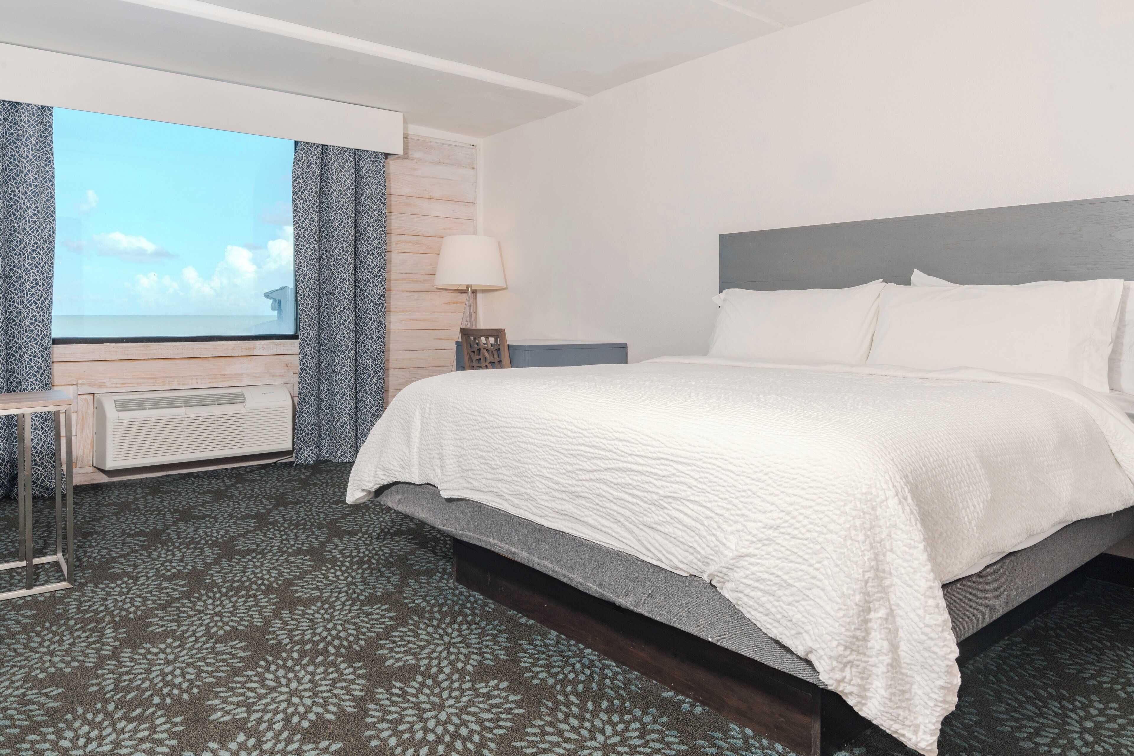 Quarto de hotel com uma cama grande, janela com vista, escrivaninha e luminária.