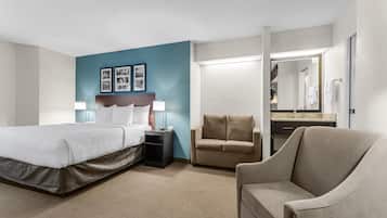 Quarto de hotel com cama, mesa de cabeceira, luminária, um sofá, uma poltrona e um banheiro visível através de uma porta aberta.