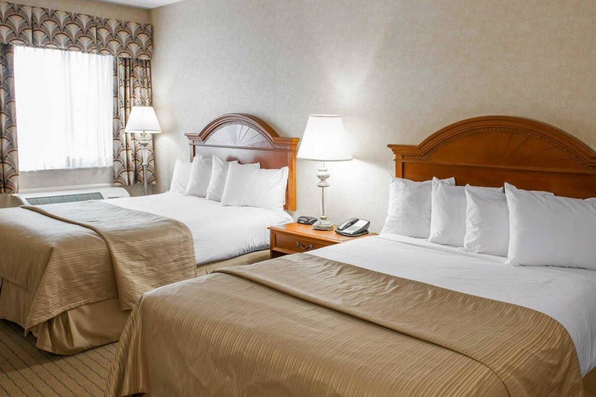 Quarto de hotel com duas camas, cada uma com cabeceira de madeira, roupa de cama branca e mesa de cabeceira com luminária e telefone.