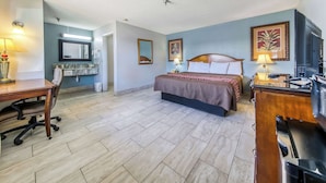 Quarto de hotel com cama, escrivaninha, cadeira e banheiro com espelho e pia.