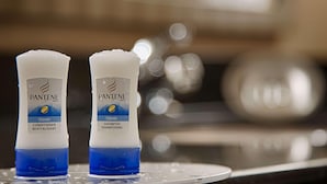 Dois produtos Pantene Pro-V Classic, um condicionador e um shampoo, sobre uma superfície refletora com fundo desfocado.