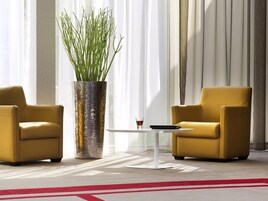 Quarto com duas poltronas amarelas, uma mesa de centro branca e uma planta grande com textura em vaso metálico.