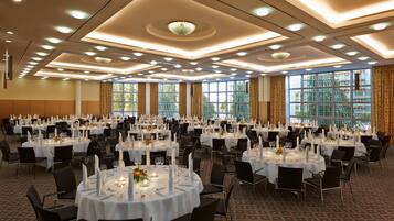 Salão de banquetes com mesas redondas preparadas para refeições, amplas janelas e teto alto com iluminação embutida.