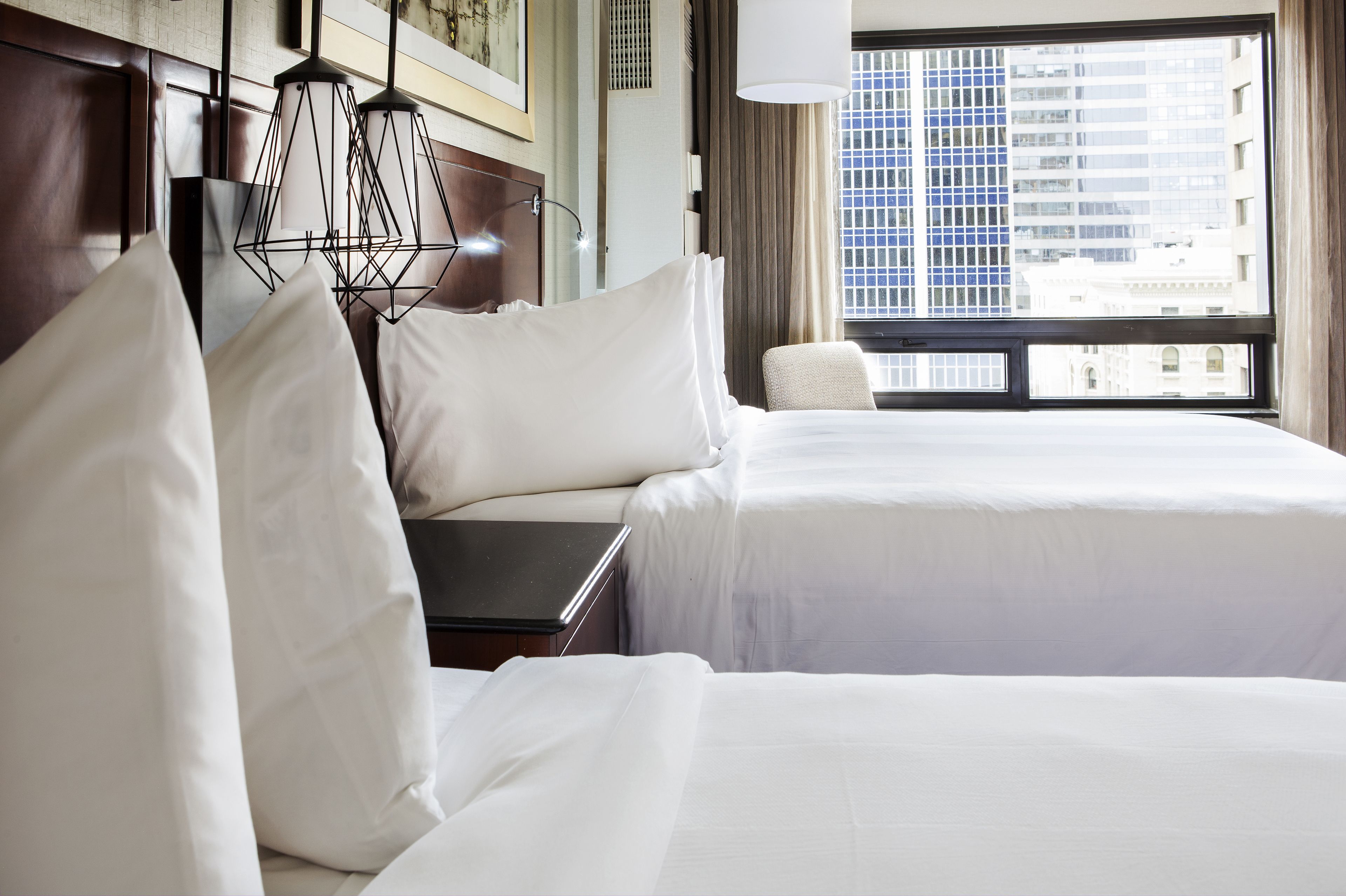 Quarto de hotel com duas camas, uma mesa de cabeceira, uma luminária e vista para o horizonte urbano por uma janela grande.