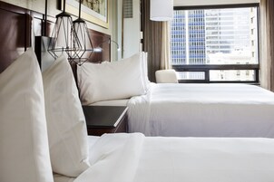 Quarto de hotel com duas camas, uma mesa de cabeceira, uma luminária e vista para o horizonte urbano por uma janela grande.