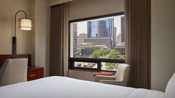 Quarto de hotel com uma janela ampla que oferece vista para a cidade, uma cama, uma poltrona, uma mesa de cabeceira e um abajur.