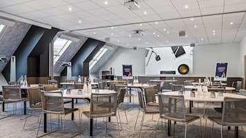 Uma sala de conferências ampla, com várias mesas e cadeiras, teto alto com claraboias e luminárias modernas.