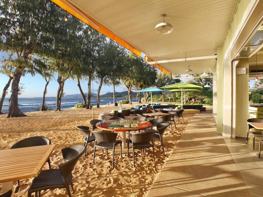 Restaurante à beira-mar com mesas e cadeiras ao ar livre, com vista para o oceano e árvores.