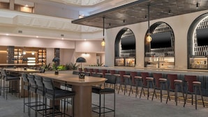 Uma área de bar moderna com uma mesa redonda central e banquetas, cercada por janelas em arco e um teto alto com padrão de grade.