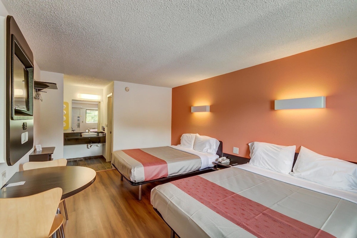 Quarto de hotel com duas camas, uma mesinha, uma cadeira, banheiro com pia e espelho e teto com acabamento texturizado.