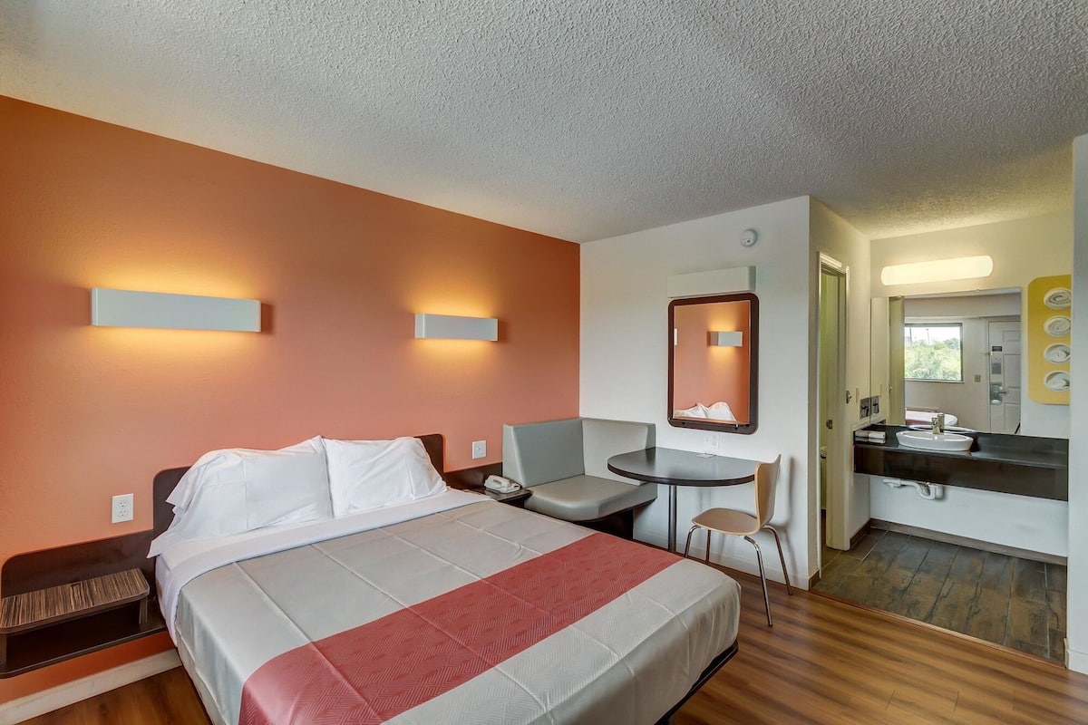 Quarto de hotel com cama, mesa de cabeceira, cadeira, escrivaninha e banheiro com pia e espelho.