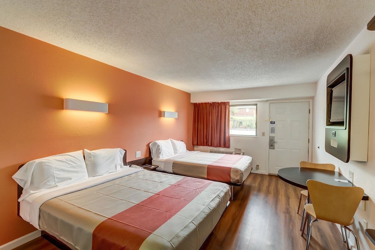 Quarto de hotel com duas camas, uma escrivaninha e uma cadeira. Há uma janela com cortinas, uma porta e uma televisão na parede.