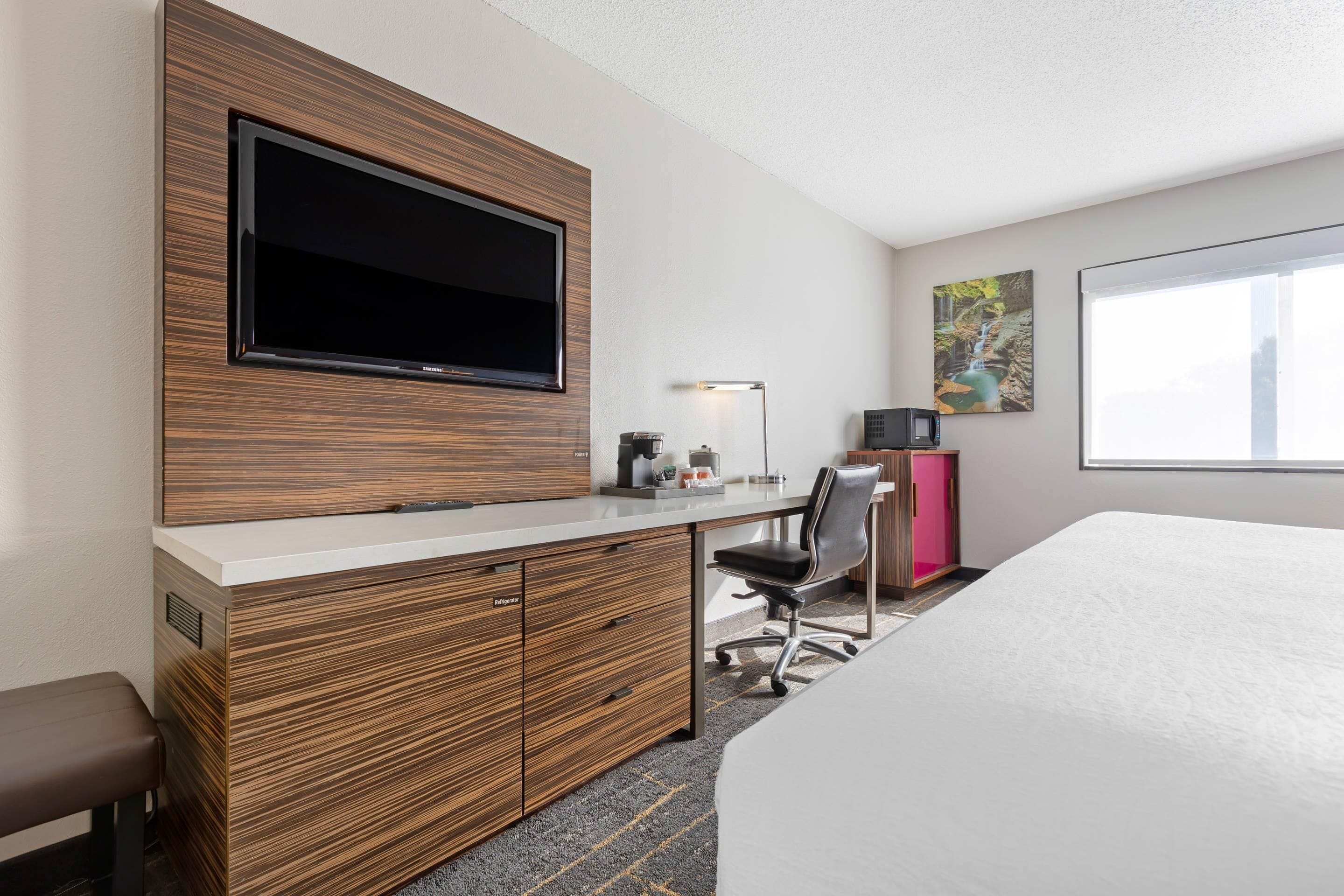 Quarto de hotel com uma cama grande, uma escrivaninha com televisão de tela plana, uma cadeira, máquina de café e um armário rosa.