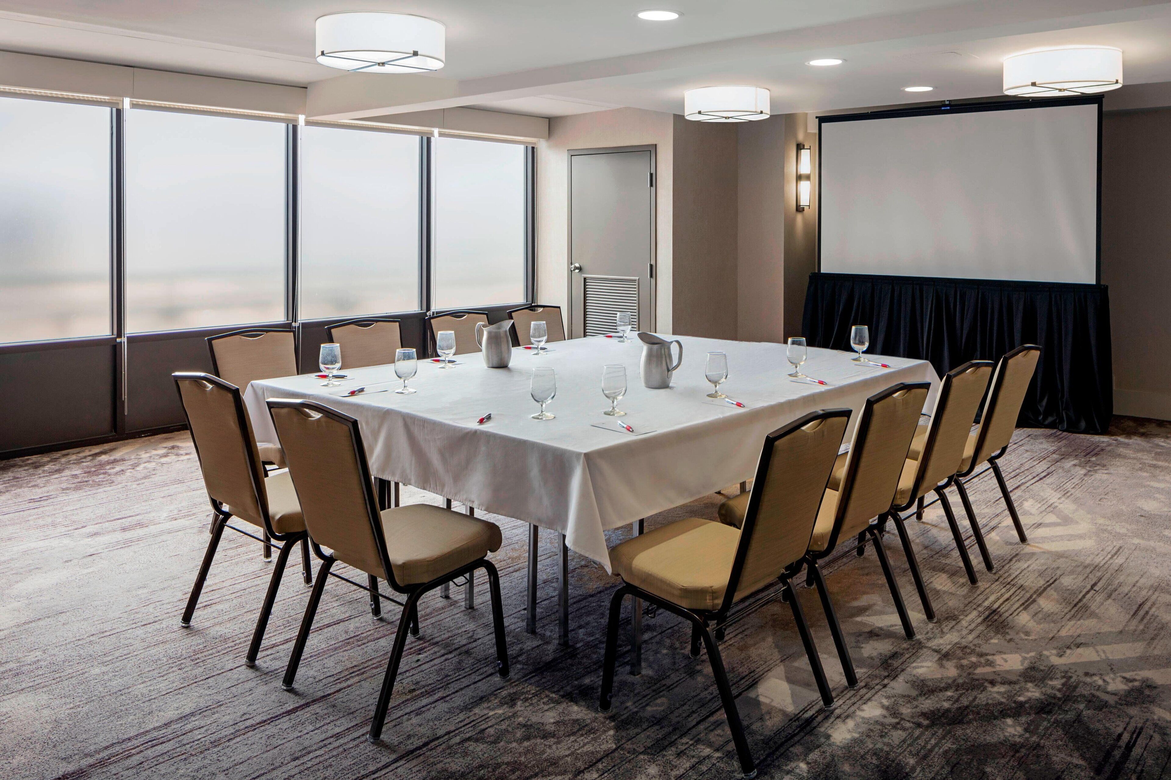 Sala de conferência com uma mesa grande, cadeiras, tela de projetor e vista através de amplas janelas.