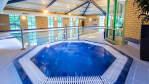 Uma grande piscina interior com hidromassagem, rodeada por teto alto com vigas de metal e amplas janelas que permitem entrada de luz natural.