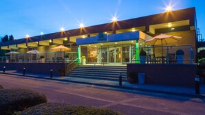 Um Holiday Inn com design moderno, apresentando fachada de vidro, área externa com mesas e guarda-sóis e uma escadaria que leva à entrada.