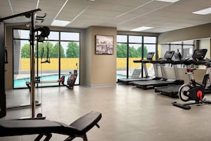 Uma academia com equipamentos de ginástica, um banco e amplas janelas com vista para uma piscina e árvores.