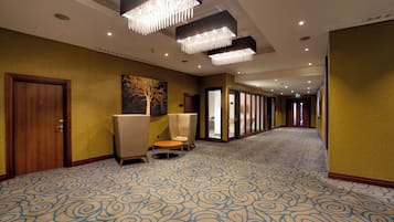 Corredor de hotel com carpete estampado, portas de madeira, área de estar com mesa redonda e lustres modernos.