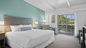 Quarto de hotel com uma cama grande, mesa de cabeceira com abajur, cadeira, pequena mesa e vista para uma varanda com piscina.