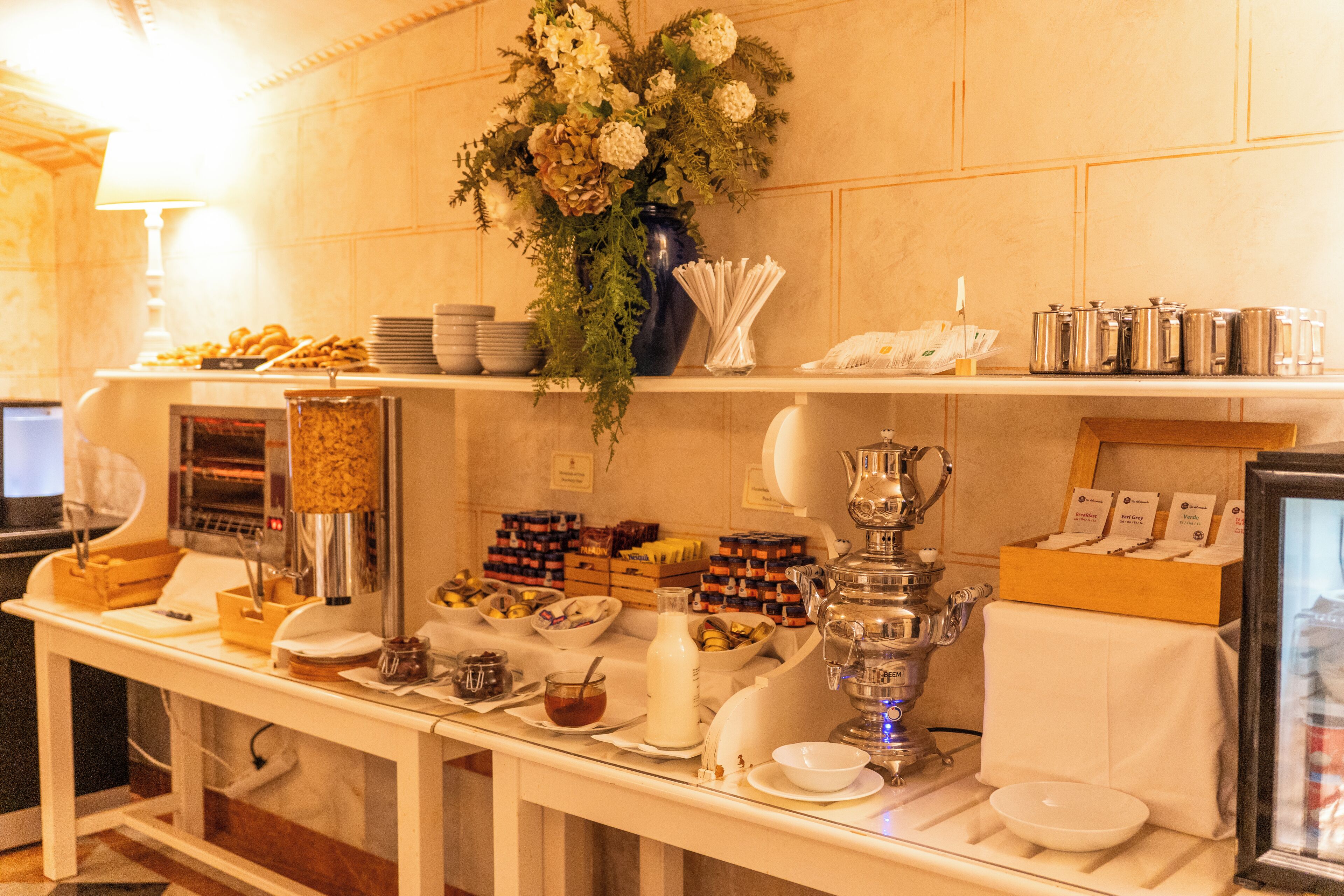 Buffet com diversos alimentos, um canto para café e um vaso decorativo com flores.