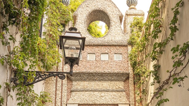 Uma ruela estreita com um arco de pedra, vasos de plantas e um lampião pendurado.