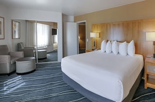 Quarto de hotel com uma cama grande, área de estar com cadeiras e um banquinho, cabeceira de madeira e vista através de uma porta.