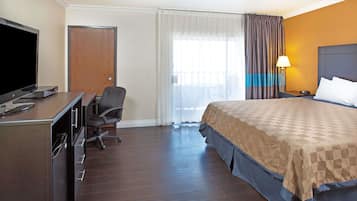 Quarto de hotel com cama, escrivaninha, televisão, cadeira, janela com cortinas e mesa de cabeceira com abajur.