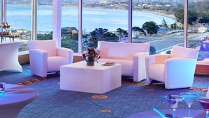Uma área de estar moderna com sofás brancos e uma mesa de centro, grandes janelas com vista para o oceano e para a cidade, e um bar com copos.