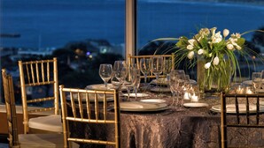 Mesa posta para refeição com cadeiras, um vaso de flores e vista para o oceano e luzes da cidade.