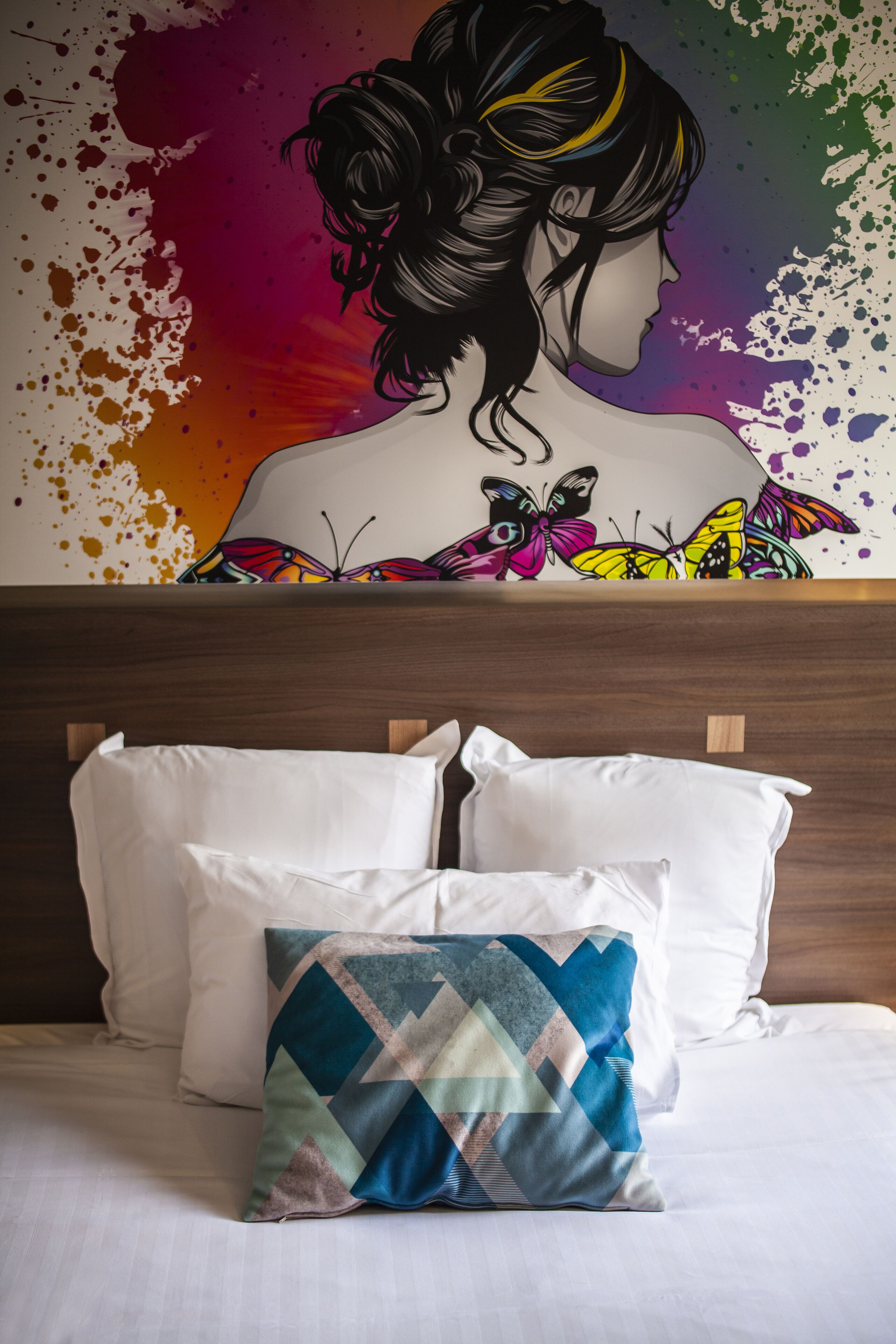 Quarto de hotel com cama, almofadas brancas, uma almofada decorativa estampada e um mural colorido na parede.