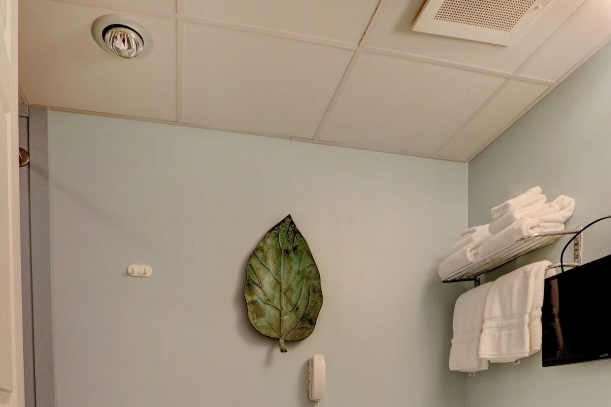 Um banheiro com vaso sanitário, pia, porta-toalhas, lixeira e uma folha decorativa na parede.