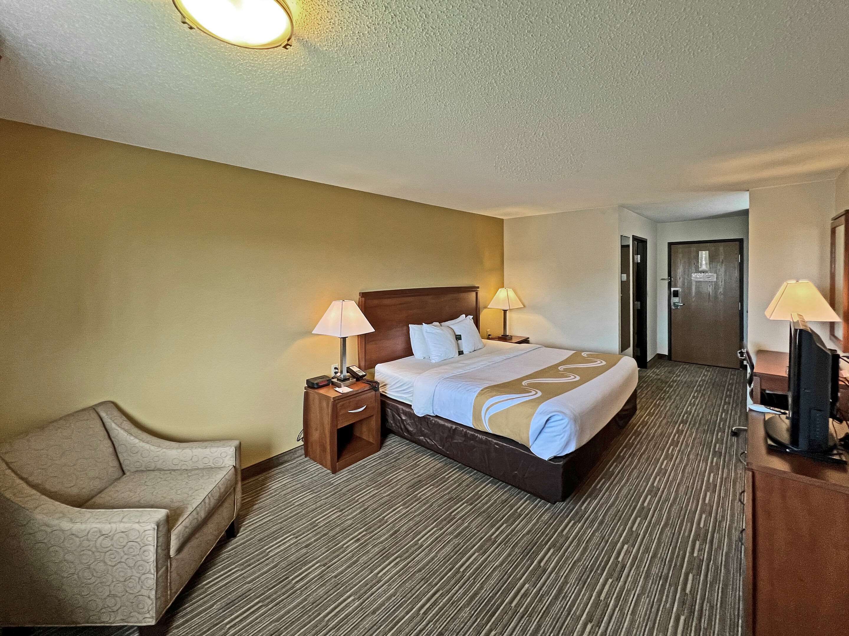 Quarto de hotel com uma cama, duas mesas de cabeceira com abajures, uma cadeira, uma televisão e uma porta que dá para outro quarto.