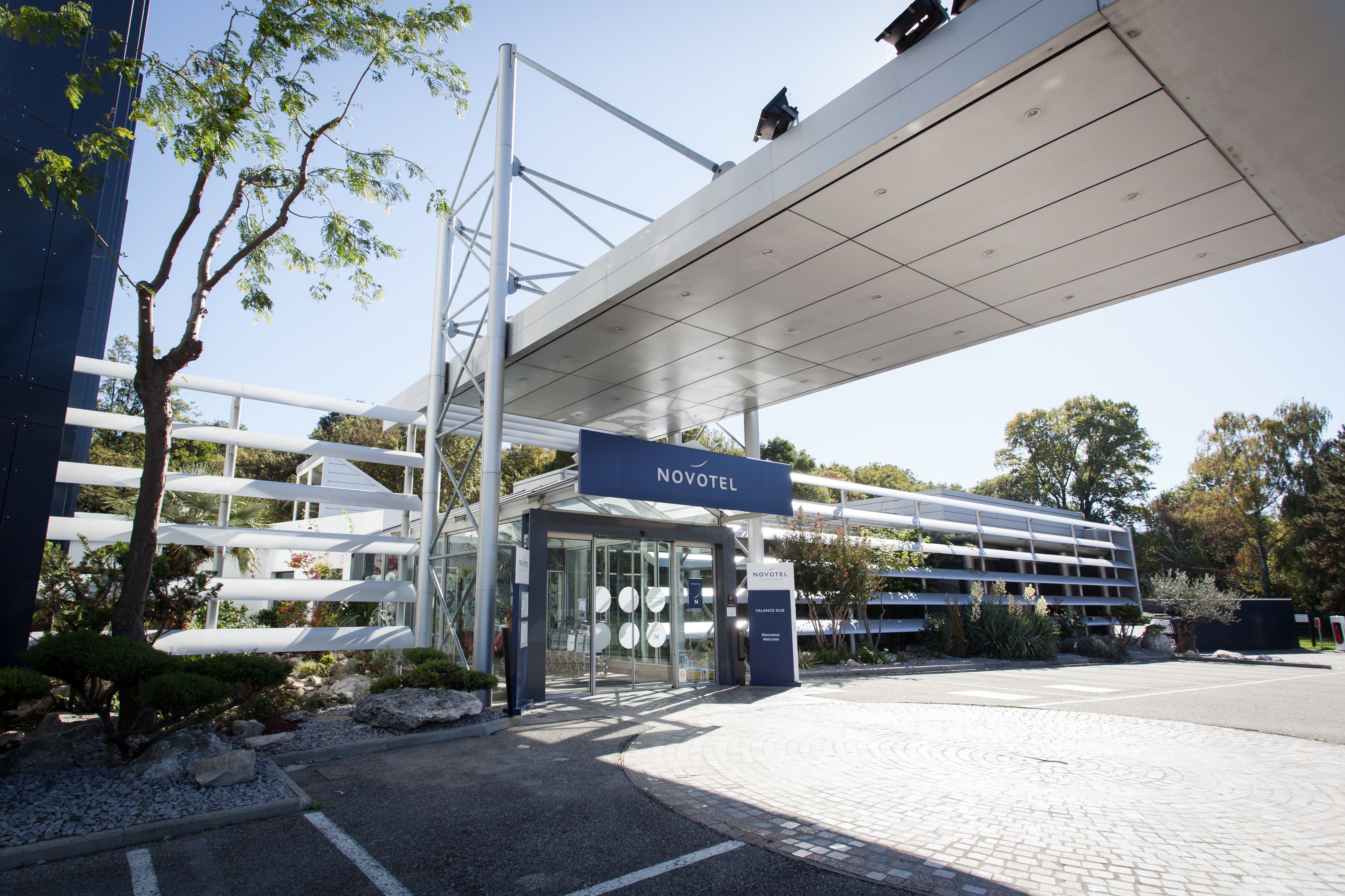 Entrada de um hotel moderno com fachada de vidro, galeria coberta e uma placa com o nome 'NOVOTEL'.