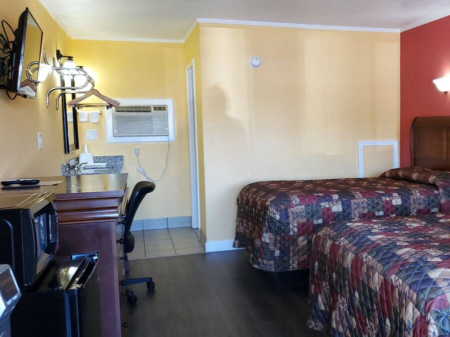 Quarto de hotel com duas camas, uma escrivaninha com cadeira, luminária de mesa, ar condicionado e luminária na parede.