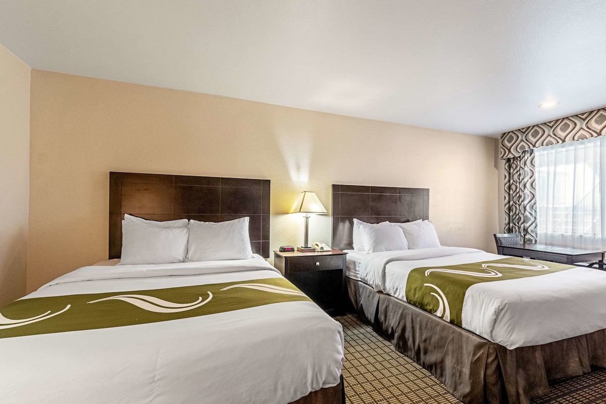 Quarto de hotel com duas camas, uma escrivaninha, uma luminária e cortina estampada.