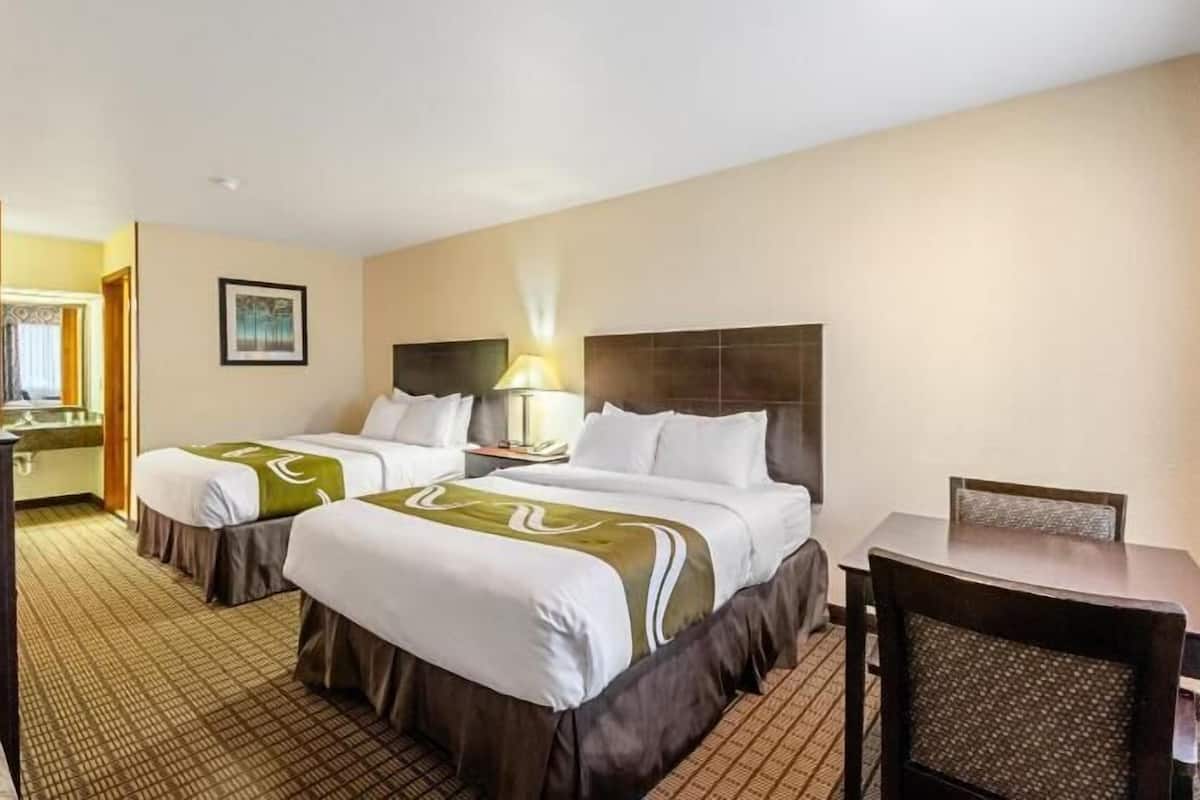 Quarto de hotel com duas camas, uma escrivaninha, uma cadeira, uma televisão e um banheiro visível ao fundo.