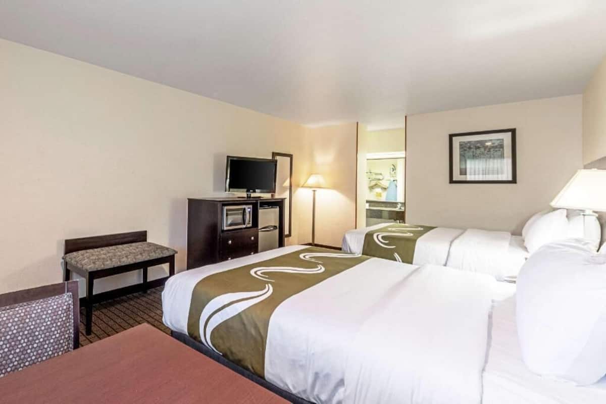 Quarto de hotel com duas camas, uma escrivaninha com televisão, uma cadeira e um abajur.
