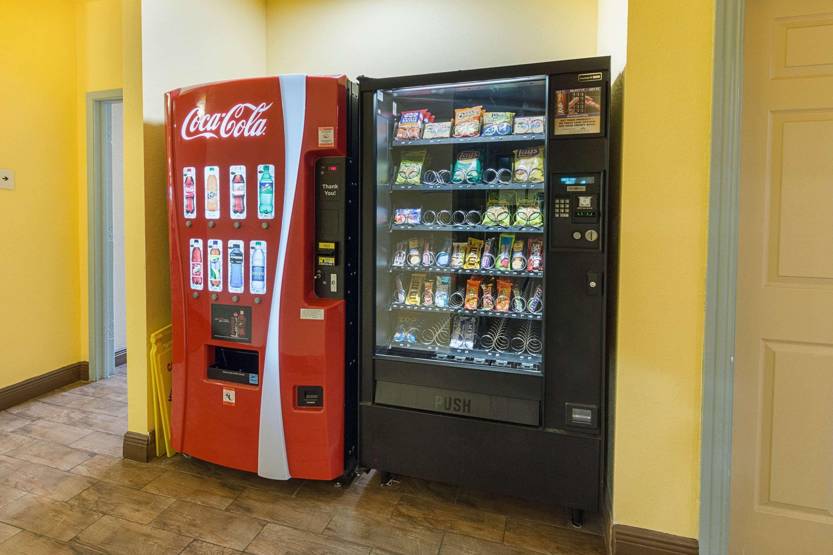 Máquina de vendas com o logotipo da Coca-Cola e interface de botões, situada em um corredor com paredes amarelas e piso de madeira.