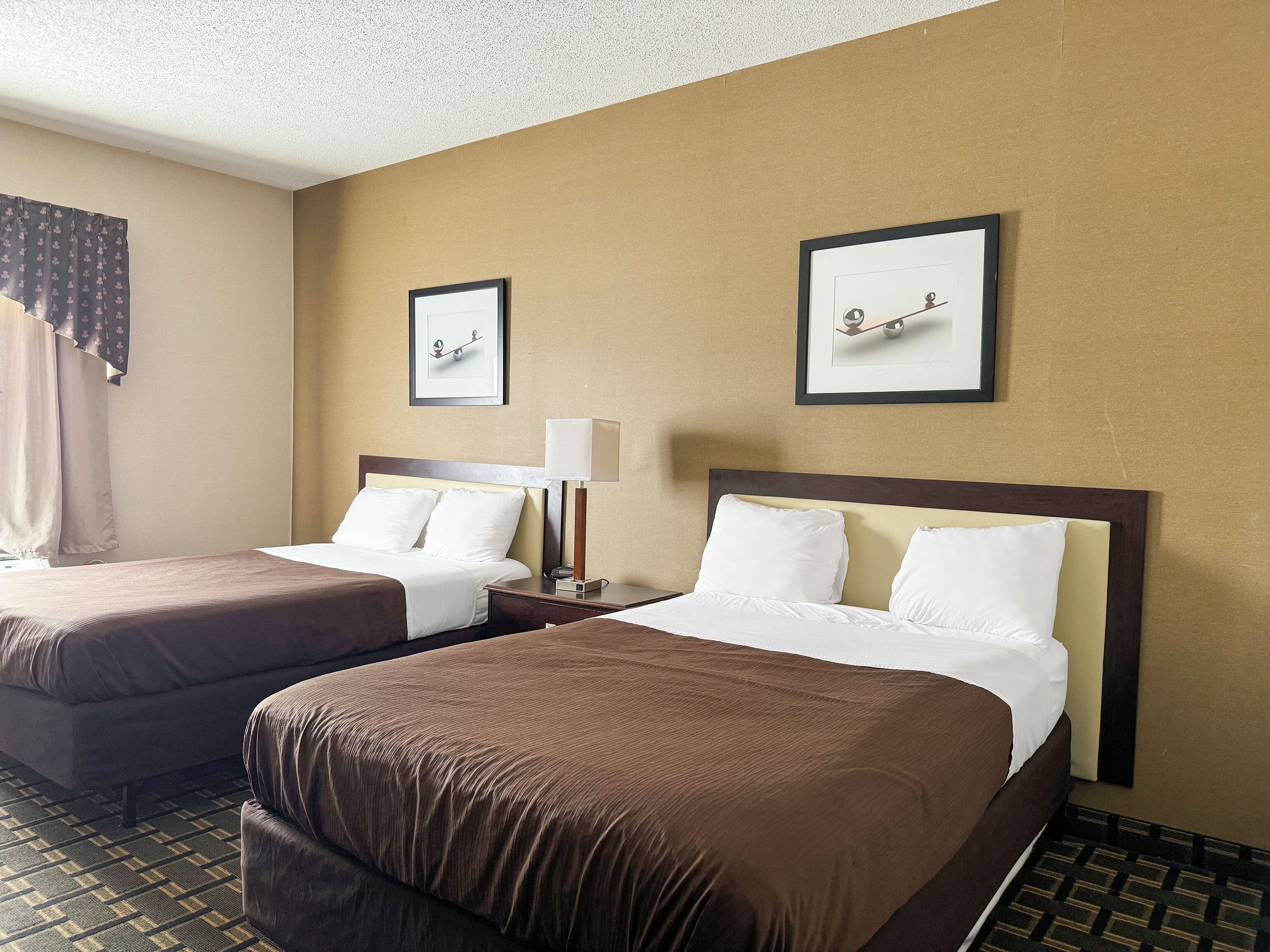 Quarto de hotel com duas camas, mesa de cabeceira com abajur e quadros nas paredes.