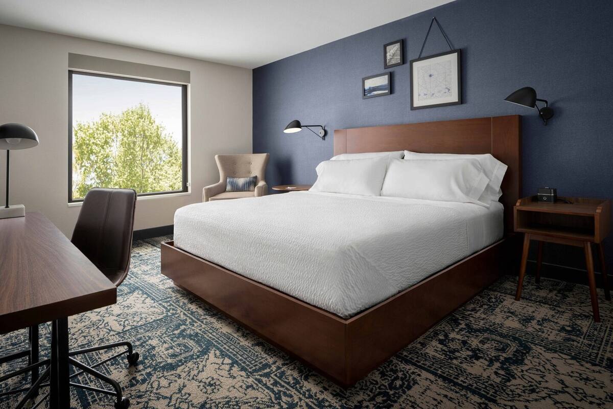 Quarto de hotel moderno com uma cama grande, uma mesa, uma cadeira e uma janela com vista para árvores.