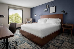 Quarto de hotel moderno com uma cama grande, uma mesa, uma cadeira e uma janela com vista para árvores.